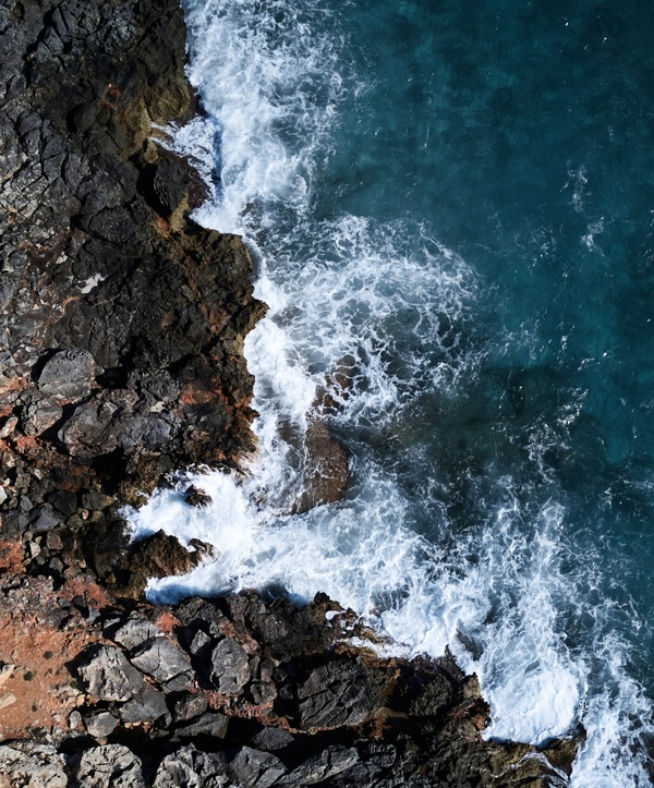 Waves crash against a rugged rocky coastline, viewed from above.