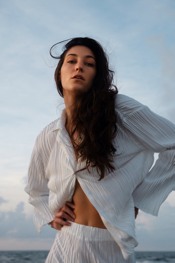 A woman in a white outfit poses with a confident expression, the ocean breeze in her hair.